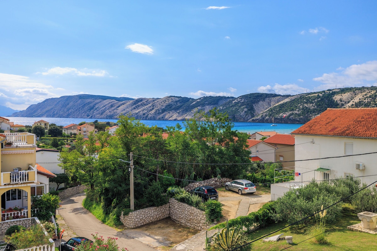 Uma área residencial costeira com casas, um muro de pedra e uma vista clara do mar e montanhas ao fundo.