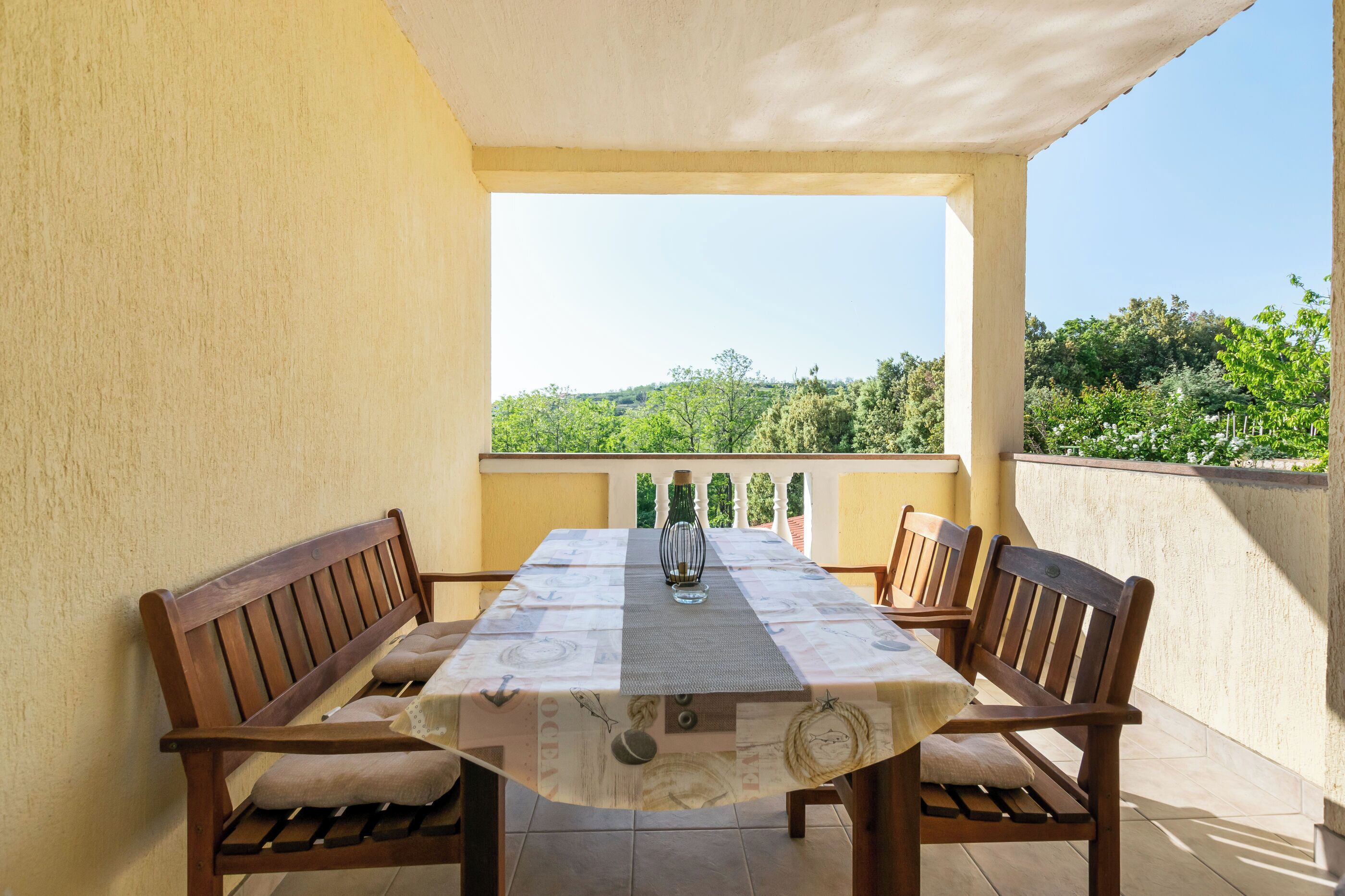 Alpendre coberto com mesa e quatro cadeiras de madeira, um vaso decorativo e vista para o verde.