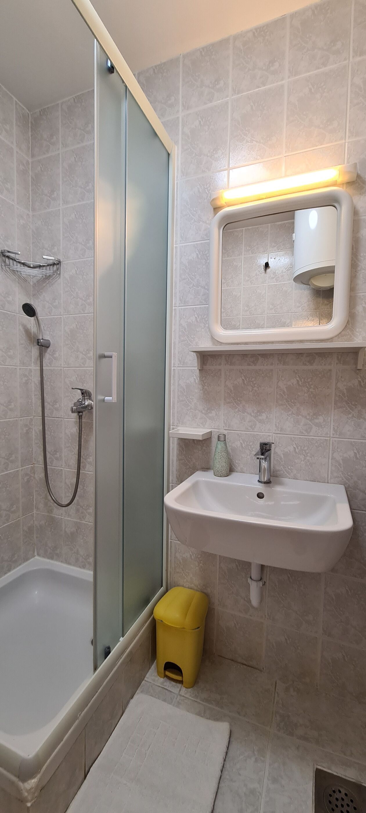 Casa de banho com cabine de duche em vidro, lavatório branco e um contentor de lixo amarelo.