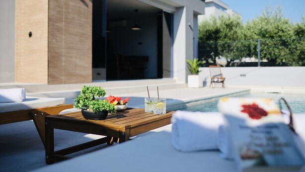 Uma área moderna com piscina ao ar livre, mesa de madeira, cadeiras de descanso e um pequeno vaso com planta.