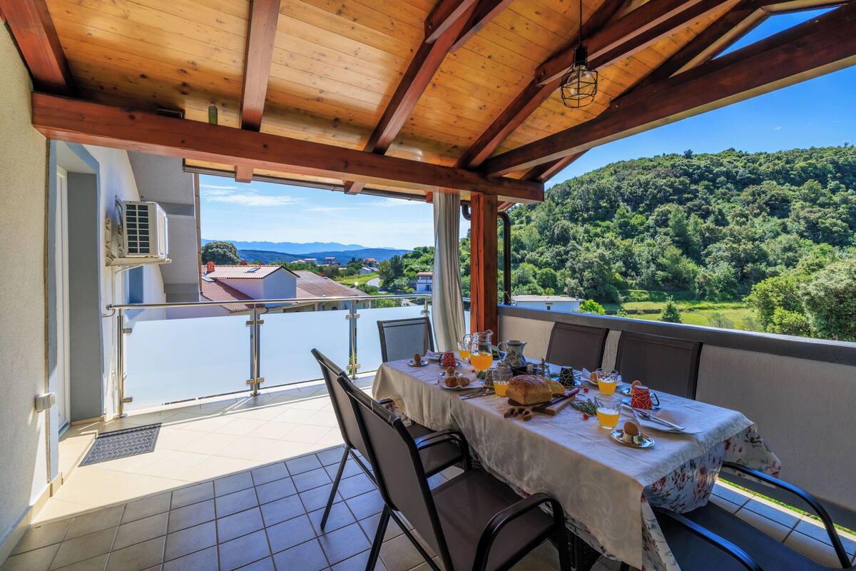 Varanda com mesa posta para o café da manhã, com vista para uma paisagem deslumbrante com colinas e um lago.