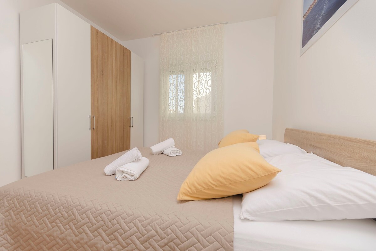 Cama bem arrumada com cabeceira acolchoada, guarda-roupa de madeira e janela com cortinas leves.