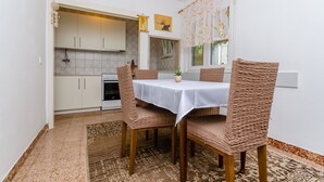 Uma sala de jantar com uma mesa posta para quatro pessoas, rodeada por cadeiras de vime, um tapete estampado e uma cozinha ao fundo.