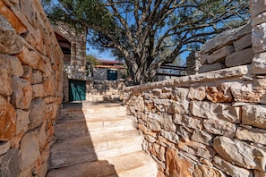 Uma escadaria de pedra leva a um edifício com uma porta verde, cercado por muros de pedra e uma grande árvore.