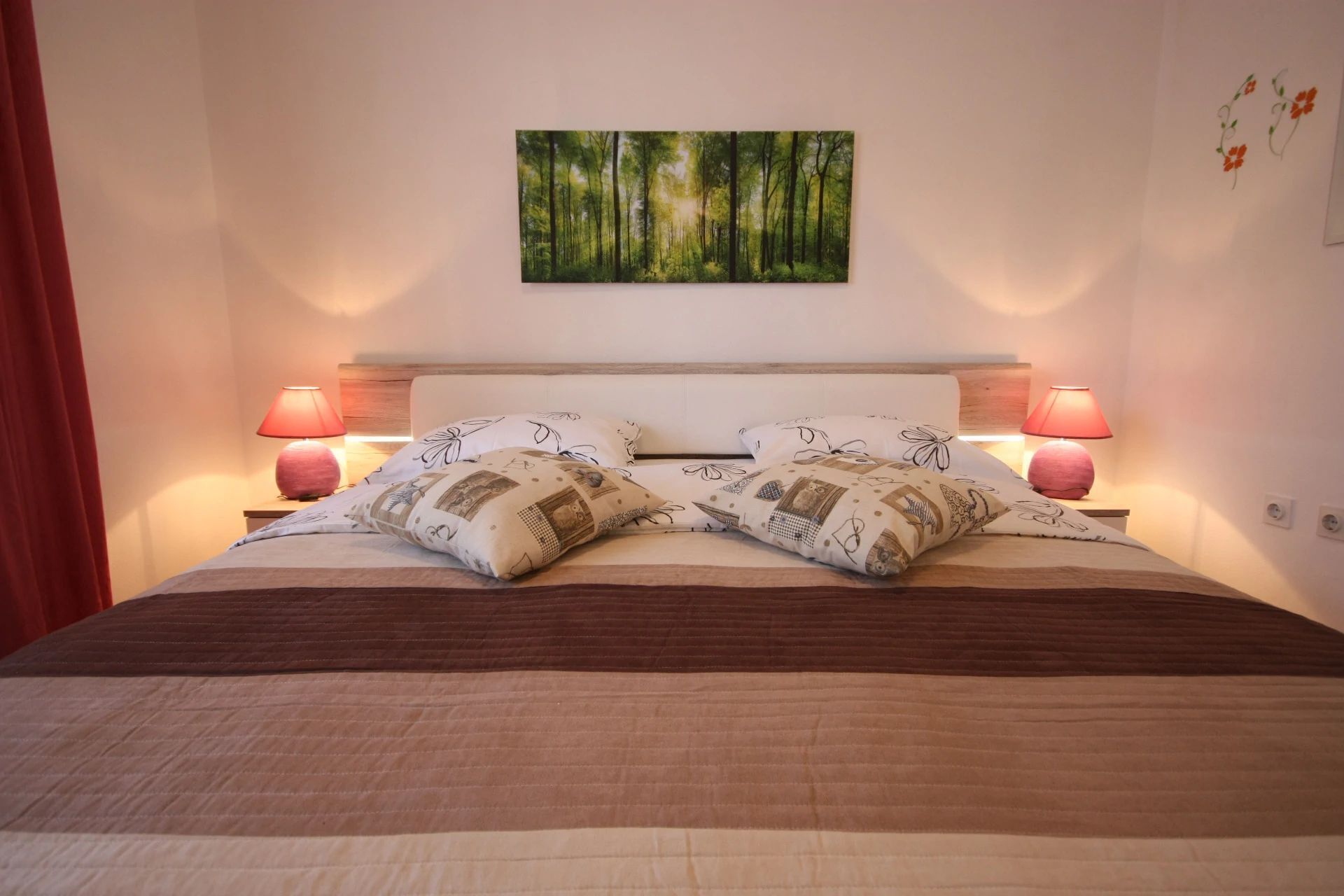 Cama bem arrumada com cabeceira marrom, duas luminárias de apoio e uma obra de arte na parede com tema de floresta.
