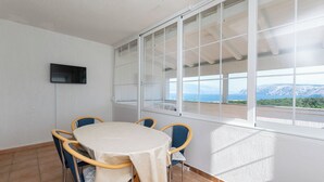 Quarto com mesa redonda, quatro cadeiras, televisão de tela plana e grandes janelas com vista para montanhas e mar.