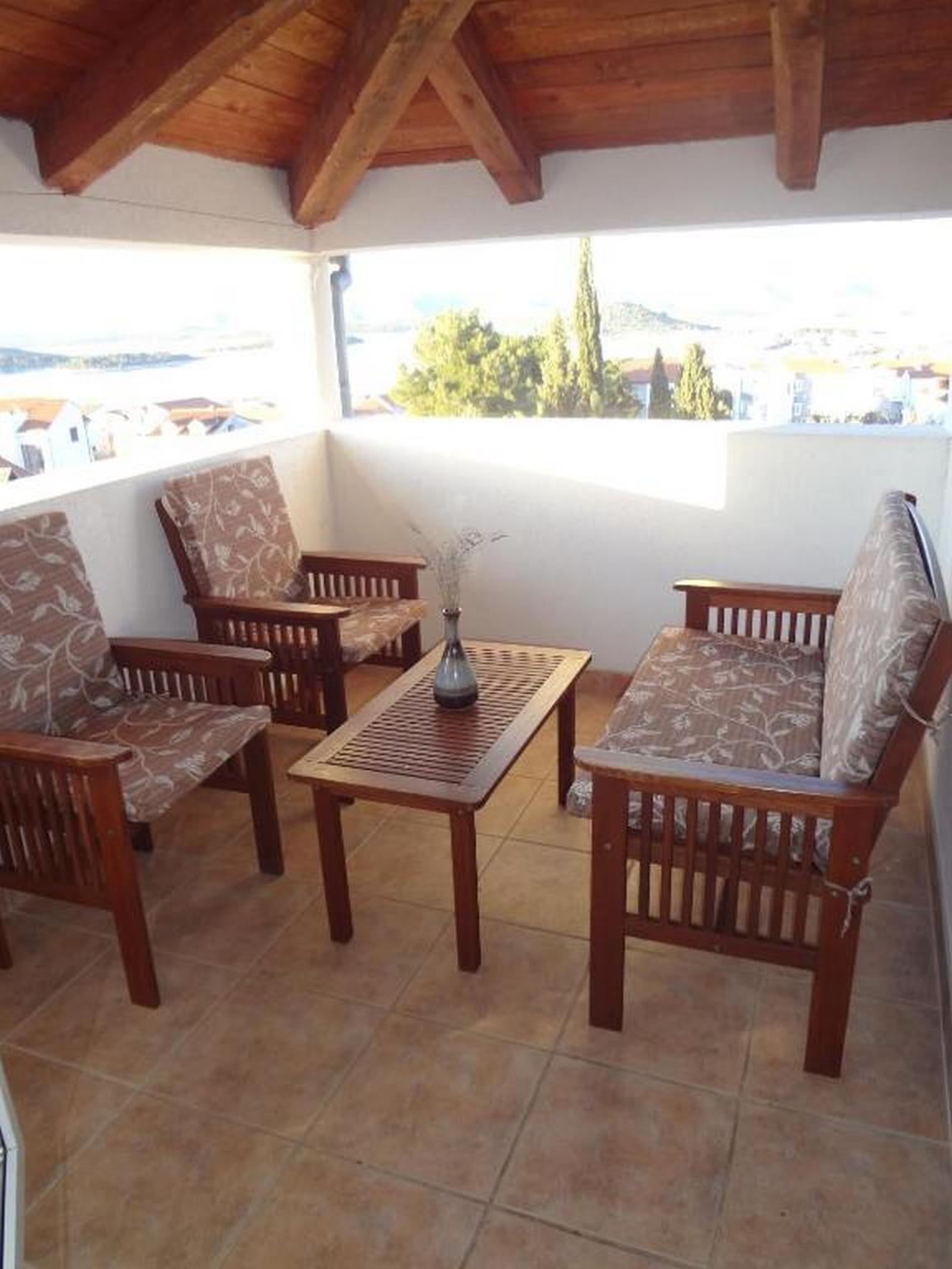 Um deck de madeira com uma mesa e quatro cadeiras, um vaso com plantas secas, e vista para edifícios e árvores.
