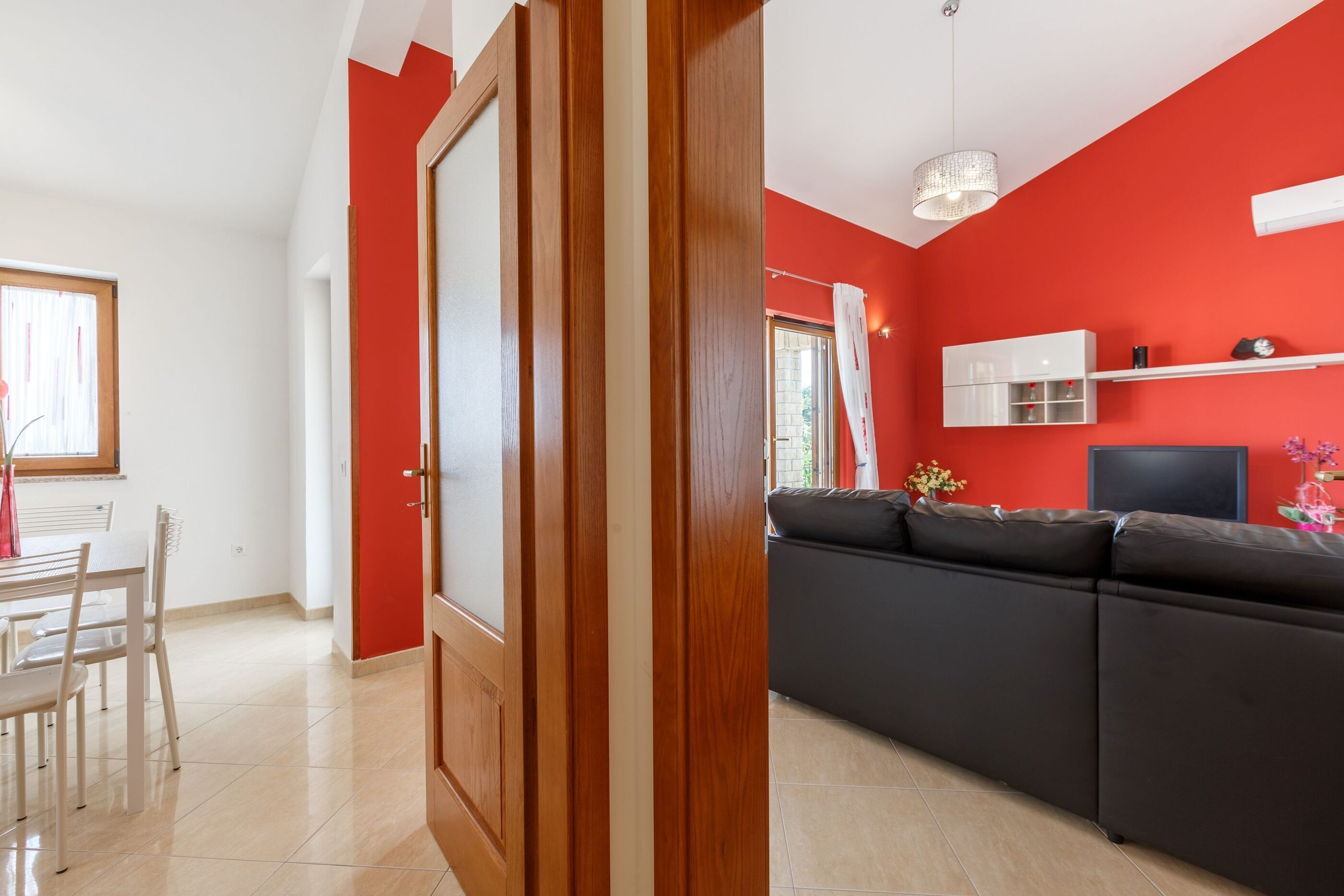 Espaço moderno para relaxar, com parede em vermelho, sofá preto, área de refeições com mesa e cadeiras, e cozinha com armário branco e televisão.