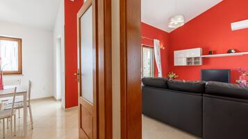 Espaço moderno para relaxar, com parede em vermelho, sofá preto, área de refeições com mesa e cadeiras, e cozinha com armário branco e televisão.