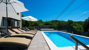 Piscina com espreguiçadeiras e guarda-sóis, cercada por uma grade e árvores.