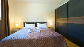 Cama bem arrumada com um edredom marrom, duas almofadas brancas e um conjunto de toalhas sobre a cama. Duas luminárias de cabeceira em ambos os lados da cama.