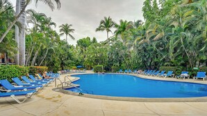 Piscina cercada por cadeiras de praia azuis, palmeiras e um edifício ao fundo.