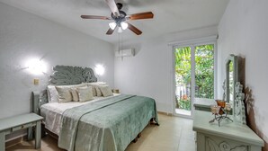 Quarto com cama, ventilador de teto, mesa de cabeceira, cômoda e janela com vista para vegetação.
