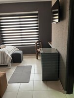 Quarto compacto com cama, uma pequena escrivaninha, uma cadeira e televisão fixada na parede.