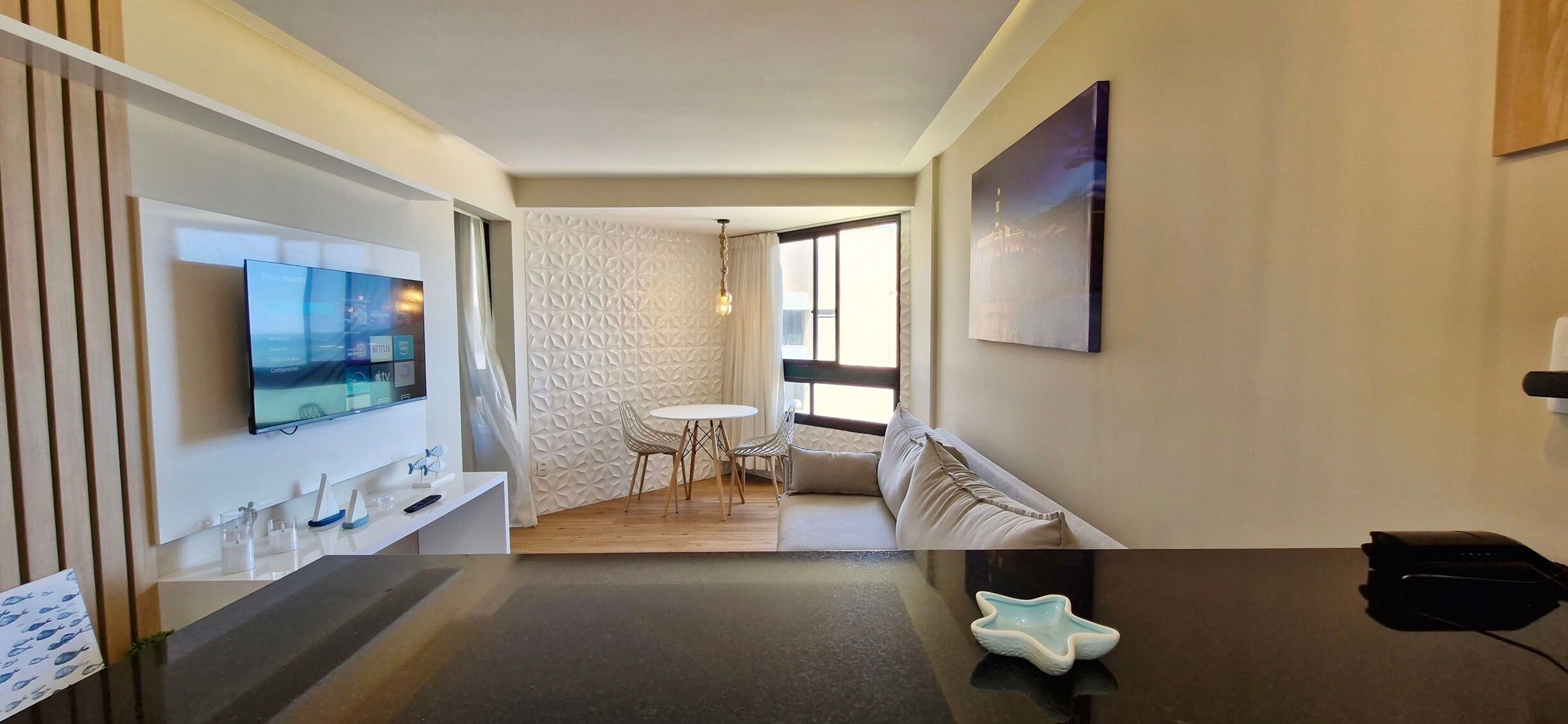 Uma sala de estar moderna com uma televisão de tela plana, uma mesa de jantar com cadeiras, um sofá e uma estrela-do-mar decorativa sobre a mesa.