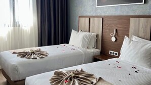 Quarto de hotel com duas camas, uma janela grande com cortinas leves, parede azul escura e uma obra de arte emoldurada sobre as camas.