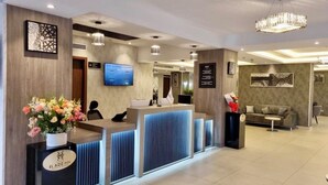 Uma área de recepção de hotel com design moderno, com balcão de atendimento, tela digital e espaço de estar com sofá e mesa pequena.