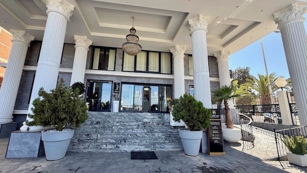 Entrada de hotel com colunas imponentes, uma escadaria majestosa e plantas em vasos.