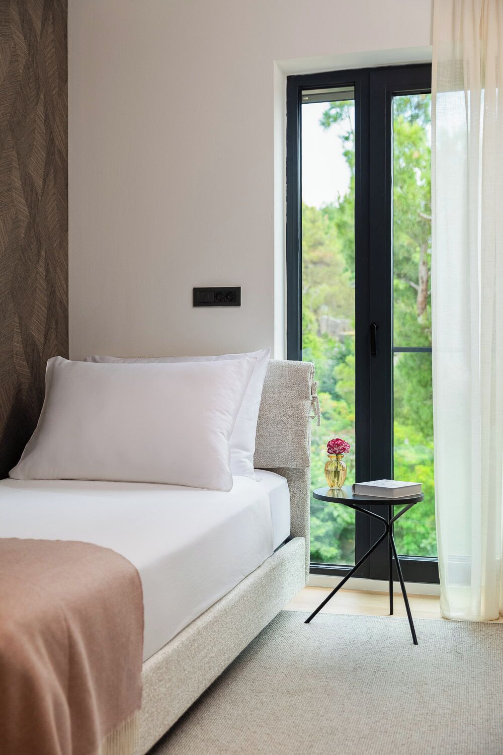 Um quarto com uma cama, uma mesa pequena com um vaso e uma janela com vista para o verde.