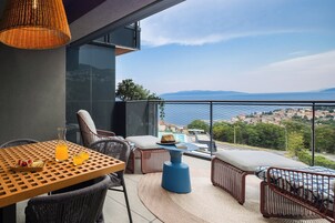 Uma varanda com móveis de vime, uma mesa com bebidas e vista para uma cidade costeira.