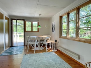 Quarto com mesa de jantar, cadeiras, luminária de chão e grandes janelas com vista para o verde.