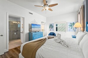 Quarto de hotel com cama, televisão, escrivaninha, cadeira, ventoinha no teto e chuveiro.