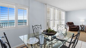 Uma área de refeições com mesa de vidro, cadeiras brancas e vista para o oceano através de persianas brancas.