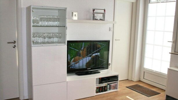 Quarto com um armário branco para entretenimento, uma televisão e um aparador de vidro com copos de vinho.