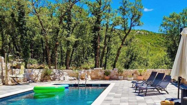 Piscina com espreguiçadeiras e uma cadeira, rodeada por árvores e um muro de pedra.