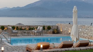 Área da piscina com espreguiçadeiras, um muro de pedra e vista para o mar e montanhas.