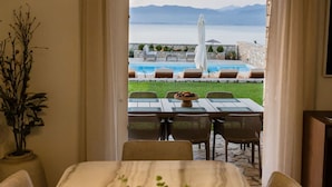 Uma área de refeições com uma mesa de mármore, cadeiras e um vaso de flores. Luminárias pendem sobre a mesa. Uma vista da piscina e das montanhas é visível pela janela.