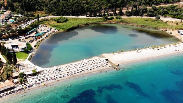 Vista aérea de uma área costeira com praia, piscina e resort com vários edifícios e guarda-sóis.