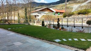 Um jardim bem cuidado com um caminho pavimentado, um pequeno lago e um edifício com telhado vermelho ao fundo.