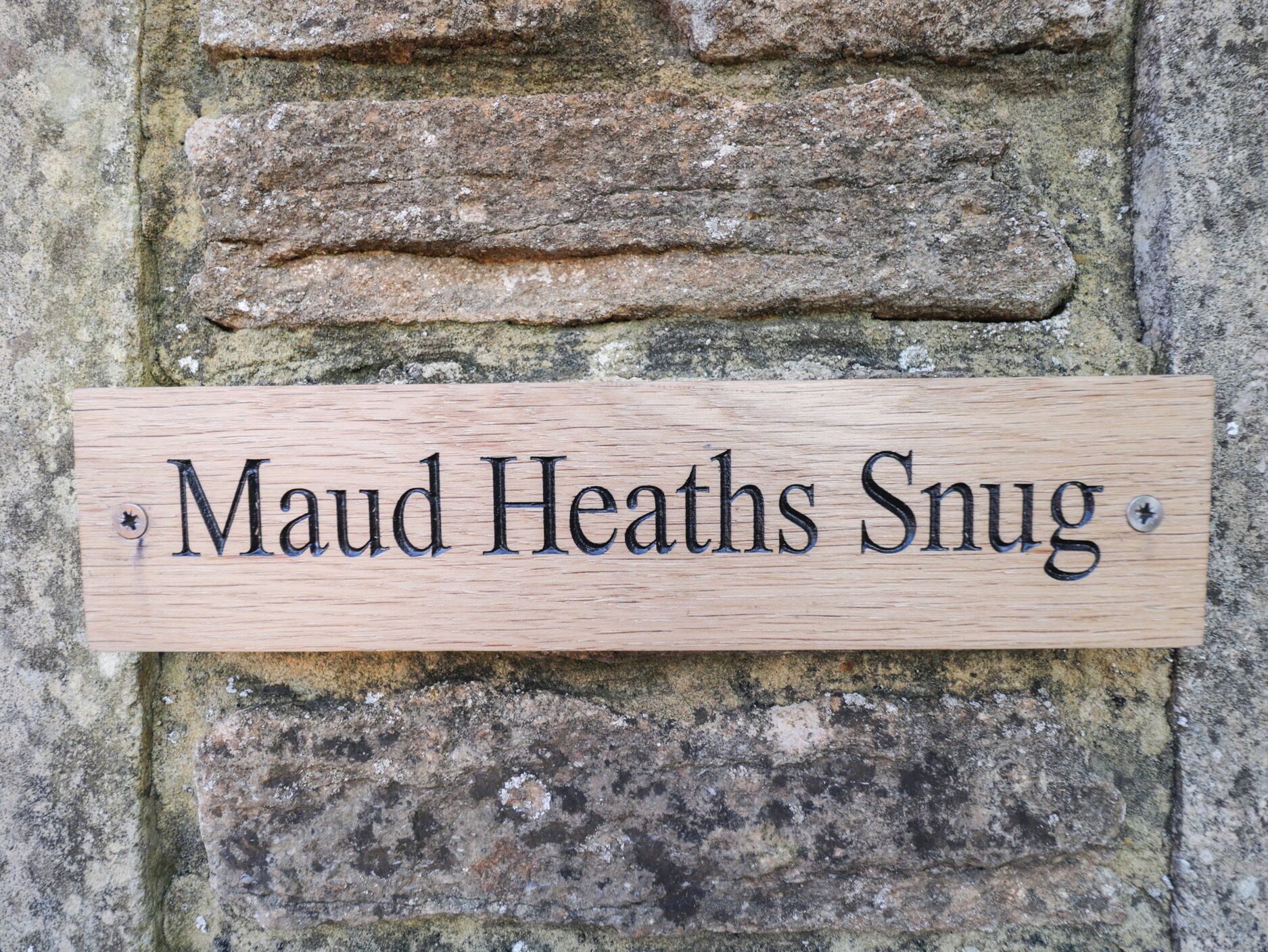 Uma placa de madeira com o texto 'Maud Heaths Snug' fixada em uma parede de pedra.