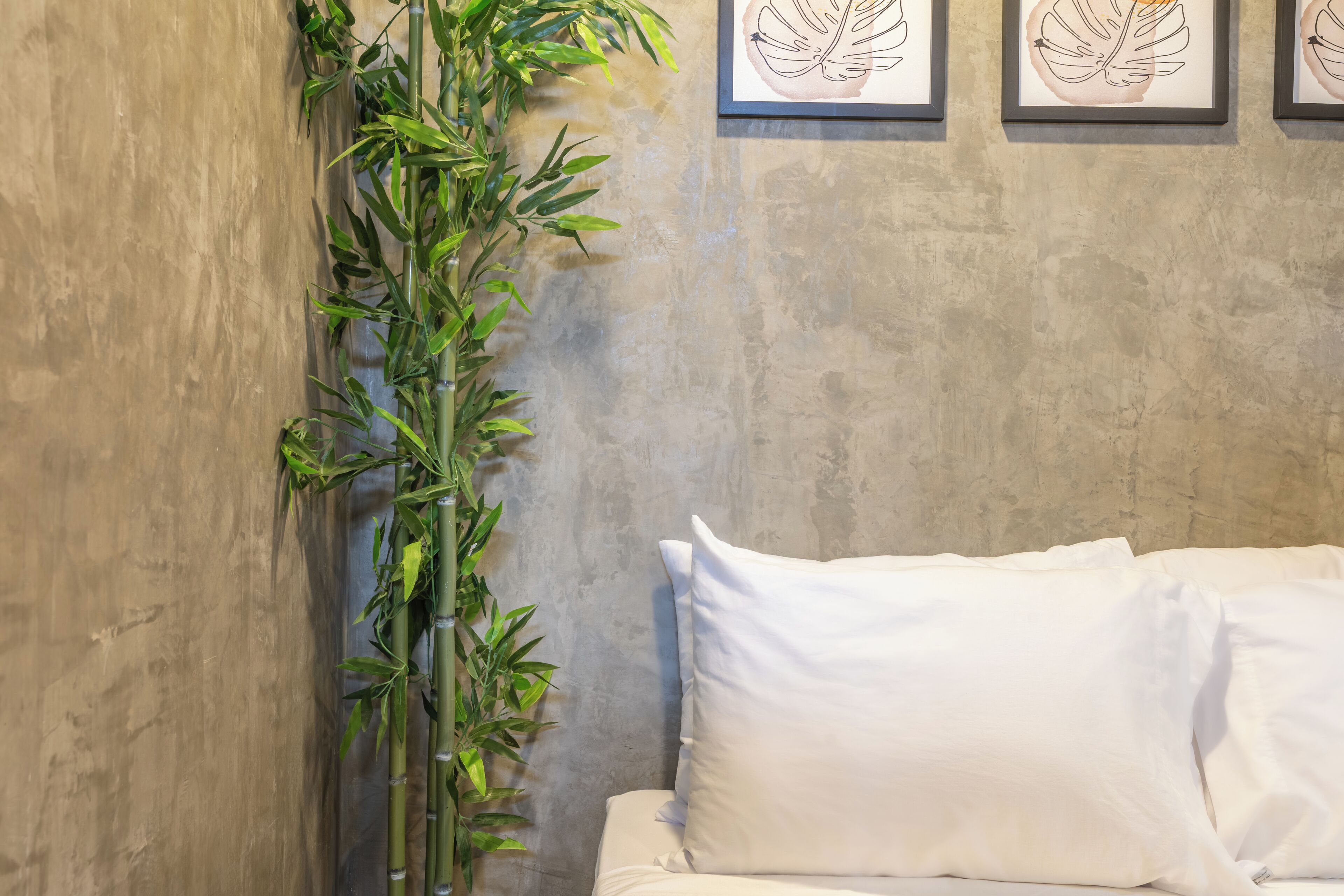 Quarto com cama, almofadas brancas, uma planta de bambu e arte botânica emoldurada na parede.