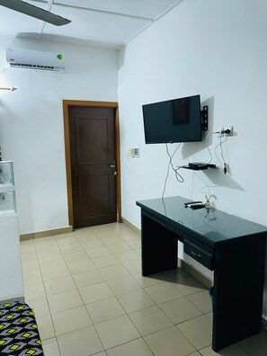 Quarto com escrivaninha, televisão fixada na parede, porta e unidade de ar condicionado.