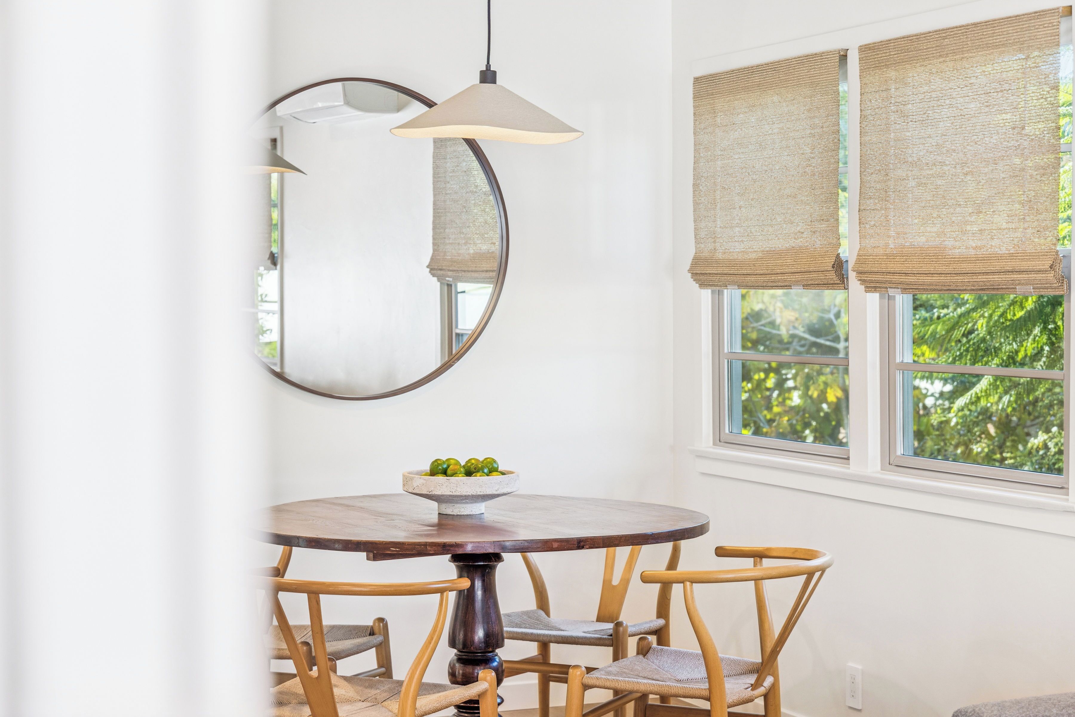 Uma área de refeições com uma mesa redonda de madeira, cadeiras de vime, uma tigela de frutas, um lustre, um espelho e uma janela com persianas.