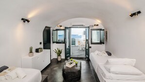 Quarto moderno e minimalista com uma grande porta em arco que dá para uma varanda. O quarto possui uma cama branca, um pequeno espaço de estar com uma mesa e um vaso de planta.