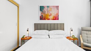 Quarto moderno com uma cama grande, duas mesas de cabeceira com abajures, uma poltrona cinza estofada e um quadro abstrato colorido na parede.