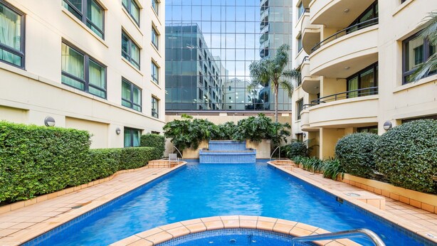 Piscina com borda azulejada, cercada por sebes e um edifício com varandas.