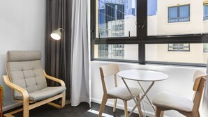 Um quarto com uma poltrona cinza estofada, uma pequena mesa redonda e um abajur. Uma janela oferece vista para edifícios lá fora.