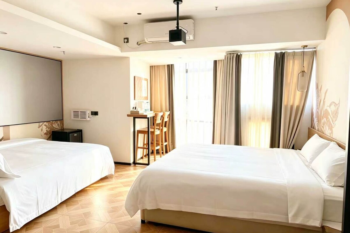 Quarto de hotel com duas camas, uma escrivaninha e uma janela grande com cortinas.