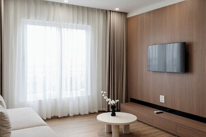Quarto de hotel moderno com uma janela grande, uma TV de tela plana na parede, uma mesa redonda com um vaso de flores e um sofá.
