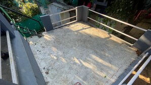 Um terraço no telhado com piso de concreto, grades de metal e uma estrutura verde ao fundo.