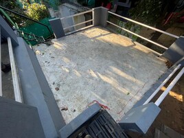 Um terraço no telhado com piso de concreto, grades de metal e uma estrutura verde ao fundo.