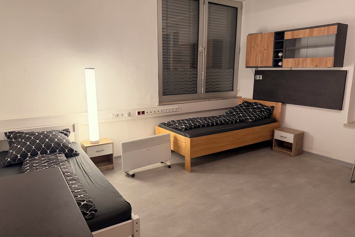 Quarto compacto com cama, escrivaninha, cadeira, janela com persianas, radiador e guarda-roupa.