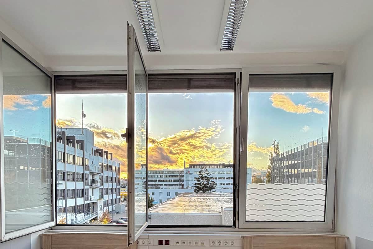 Quarto com duas camas, grandes janelas e vista para edifícios e árvores.