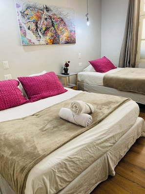 Quarto de hotel com duas camas, cada uma com colcha bege e almofadas cor-de-rosa. Há um quadro na parede, uma mesinha com uma planta em vaso e um lustre.