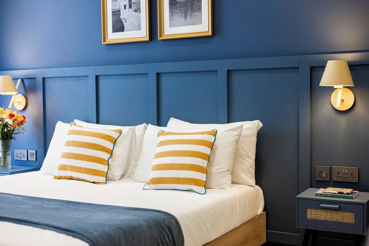 Quarto de hotel com cama, duas almofadas, mesa de cabeceira, abajur e duas fotos emolduradas na parede.