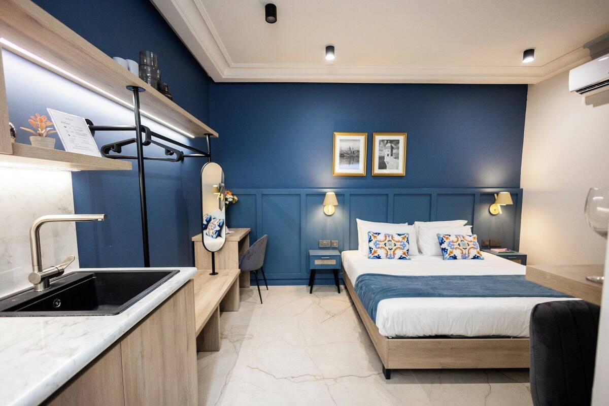 Quarto de hotel moderno com cama, mesas de cabeceira, escrivaninha com cadeira, espelho e pia embutida.